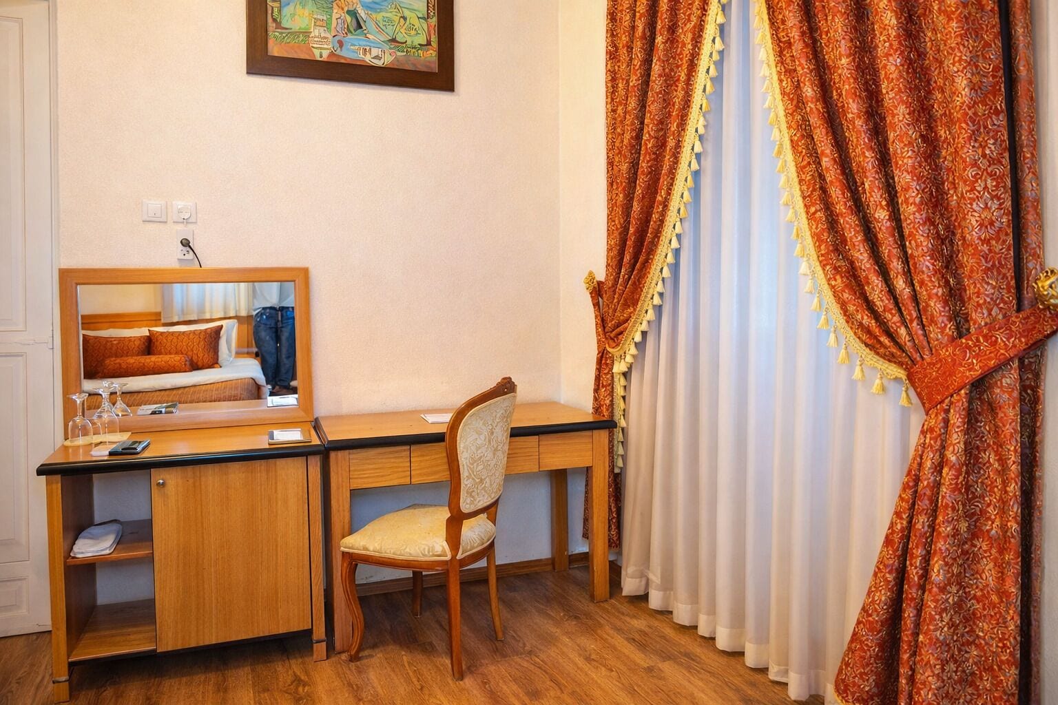 Quarto de hotel com uma escrivaninha de madeira, uma cadeira, um espelho e uma cama com roupa de cama laranja.