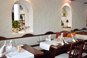 Restaurante com interior rústico, com mesas e bancos de madeira, paredes brancas e detalhes decorativos como uma escultura de pássaro e plantas penduradas.