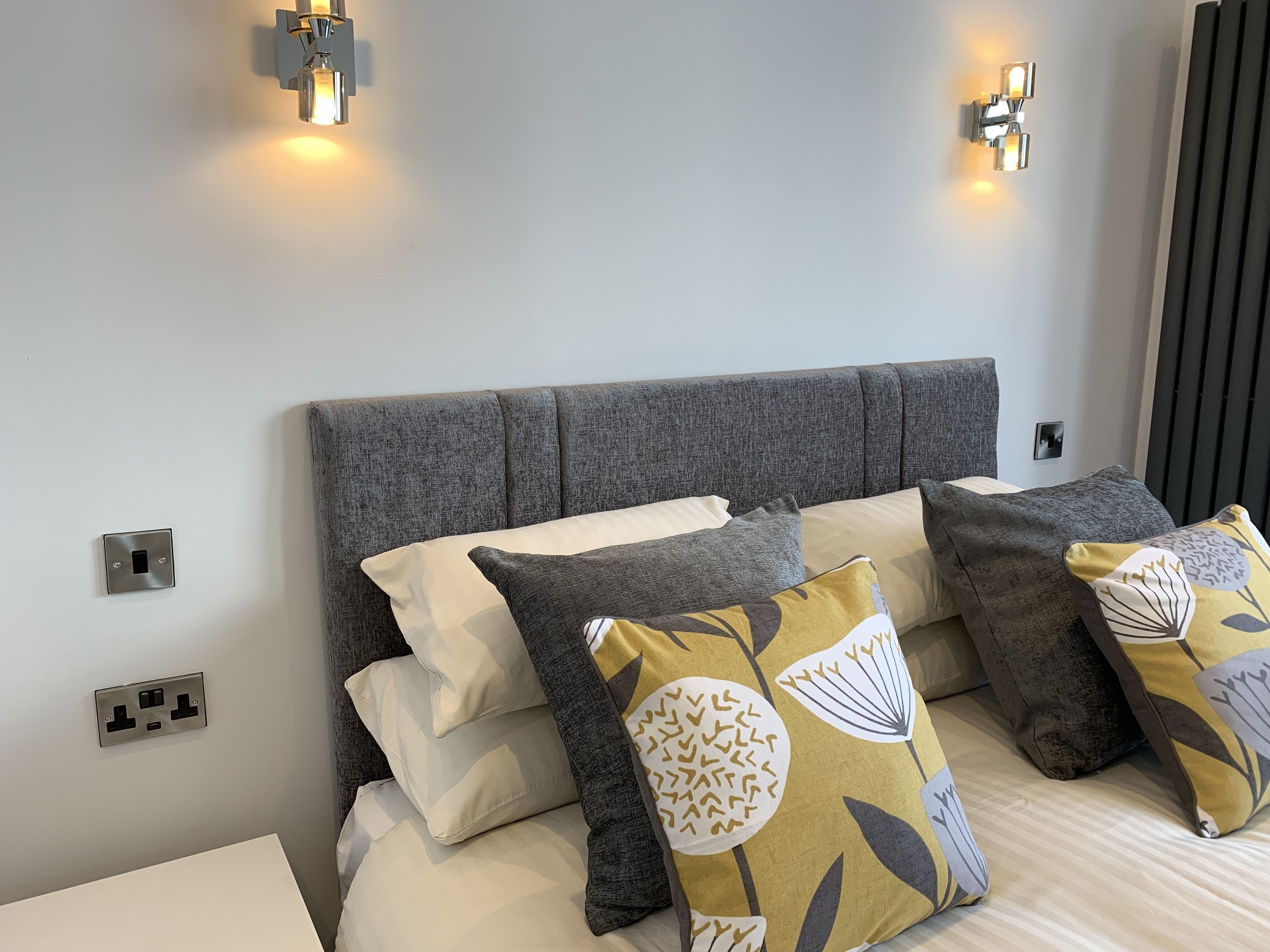 Cama bem arrumada com cabeceira cinza, várias almofadas e luminárias fixadas na parede.