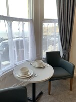 Quarto com uma mesa posta para dois, dois bowls, dois pratos e talheres. Há duas cadeiras, uma preta e outra verde escura. O quarto possui grandes janelas com cortinas leves e vista para carros estacionados e edifícios.