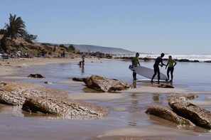 Uma praia com pessoas caminhando e carregando pranchas de surf, terreno rochoso e palmeiras.