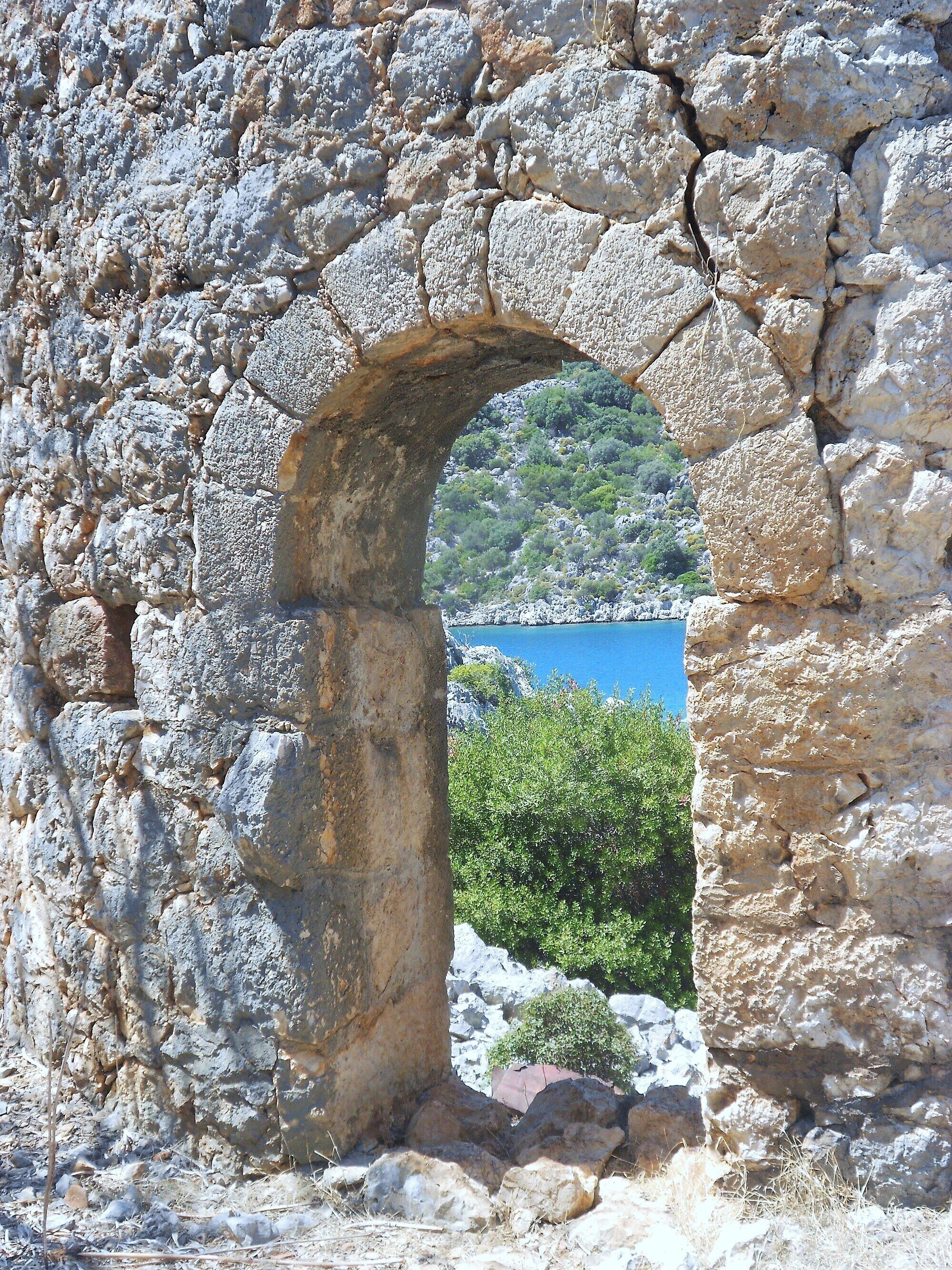 Um antigo arco de pedra com vista para um corpo de água azul e vegetação ao fundo.