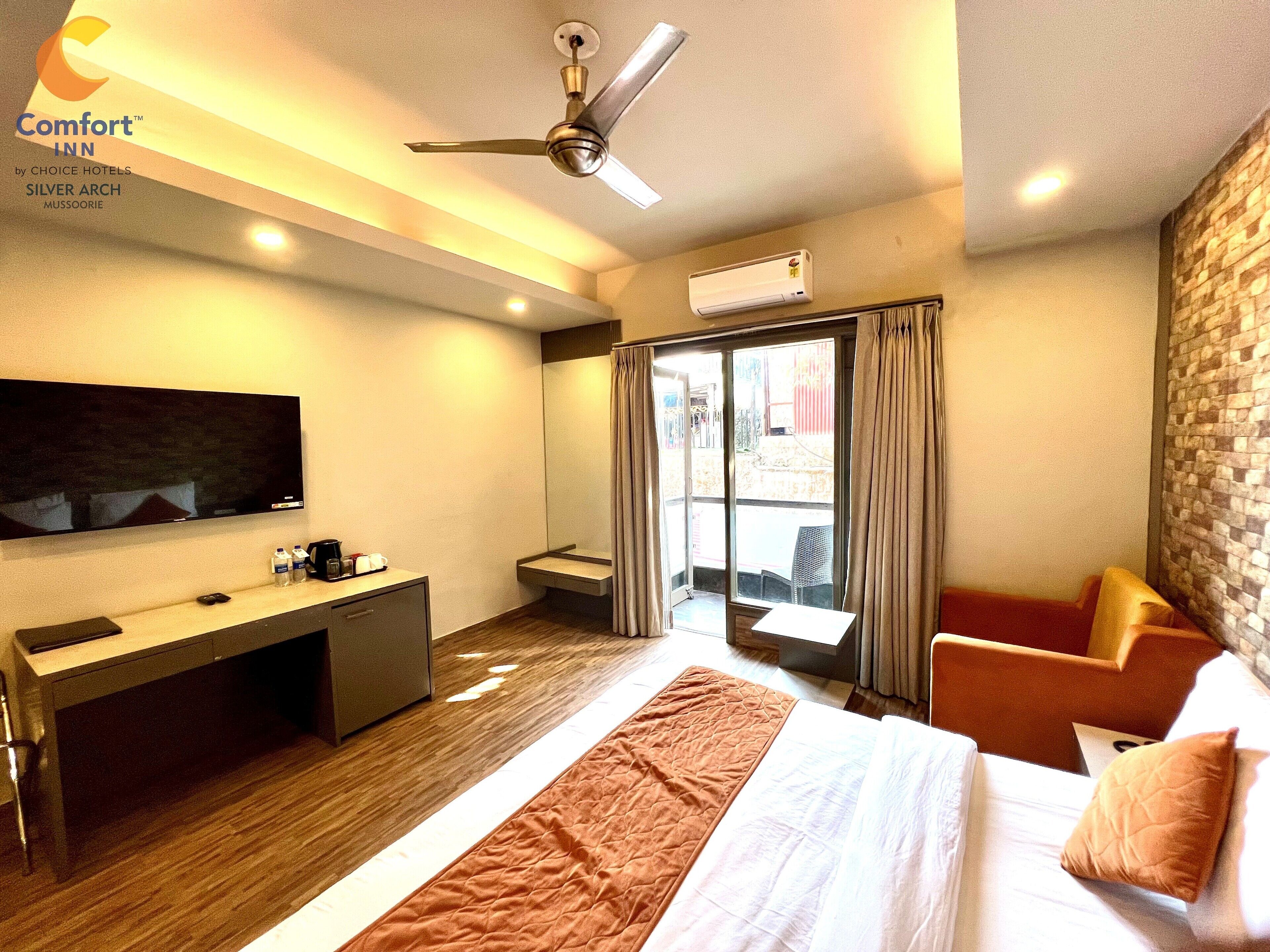 Quarto de hotel com cama, escrivaninha, TV de tela plana, ventilador de teto e uma grande janela com cortinas.