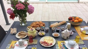 Uma mesa posta com itens de café da manhã, incluindo pão, doces, ovos e frutas, com um vaso de flores e uma revista intitulada 'CHOCOLAT MENIER'.