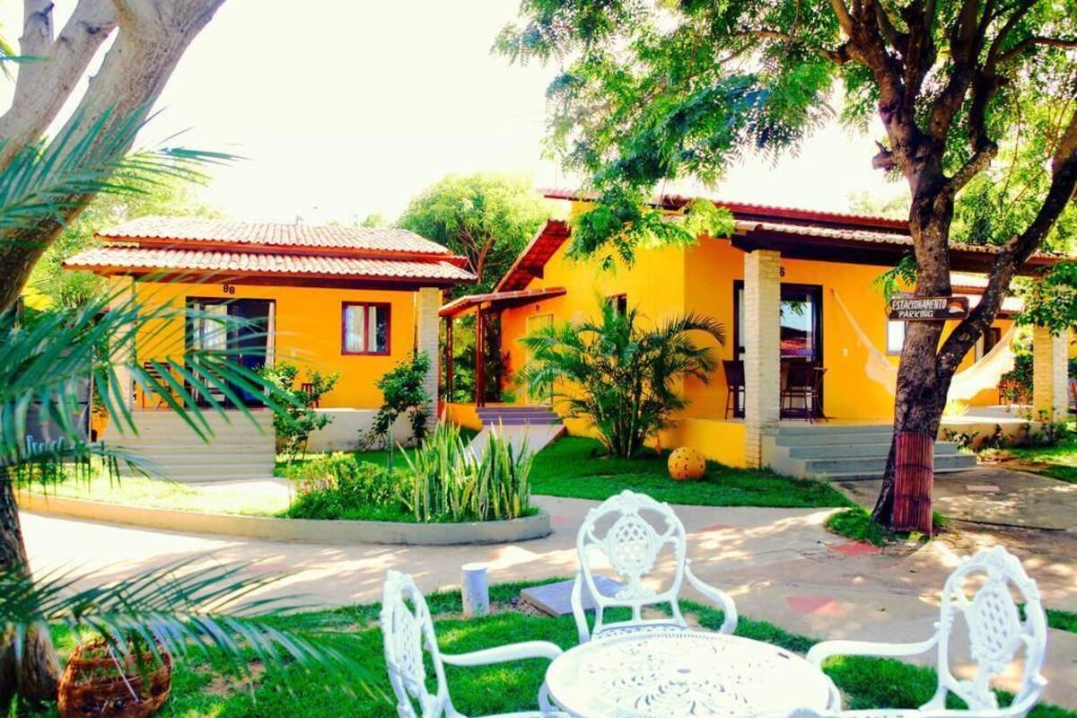 Um edifício amarelo com telhado vermelho, rodeado de vegetação e mobiliário de exterior branco.