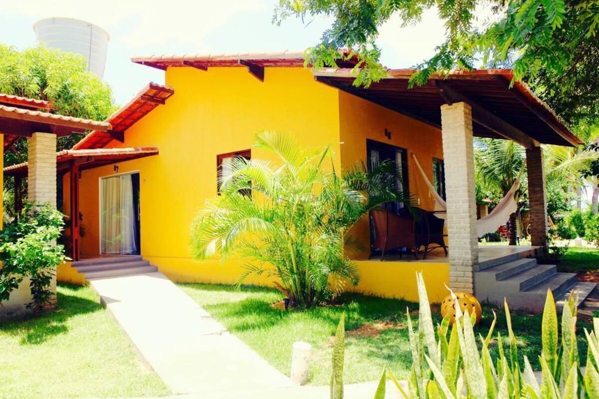 Um edifício amarelo com varanda coberta, degraus que levam à entrada e um pequeno jardim com palmeiras.