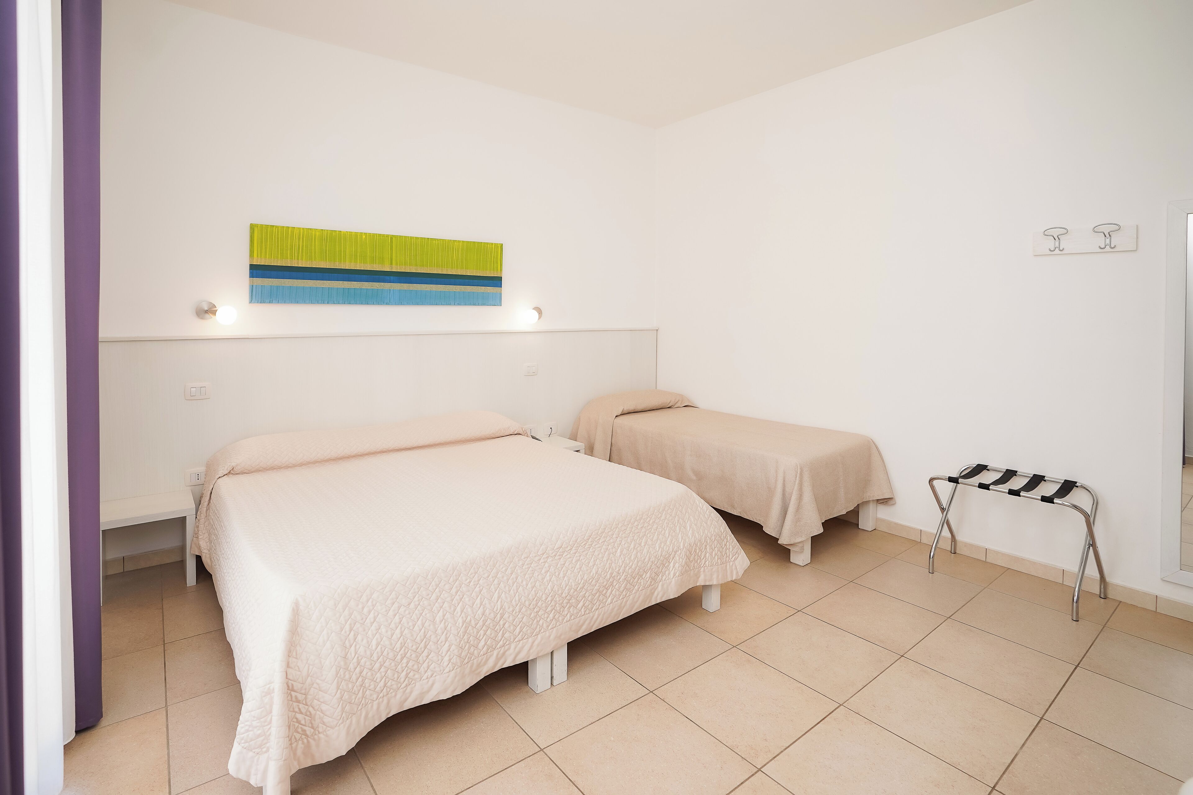 Quarto com duas camas, um banco e luminária fixada na parede.