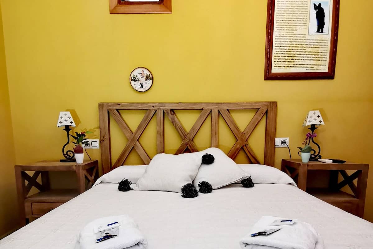 Cama arrumada com lençóis brancos e duas toalhas dobradas no pé da cama. Tem uma cabeceira de madeira, uma janela pequena e um cartaz informativo emoldurado na parede.