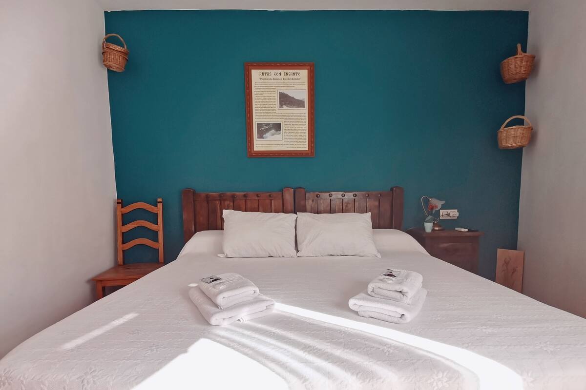 Cama bem arrumada com duas toalhas dobradas, cabeceira de madeira, mesa de cabeceira com abajur e um cartaz informativo emoldurado na parede.