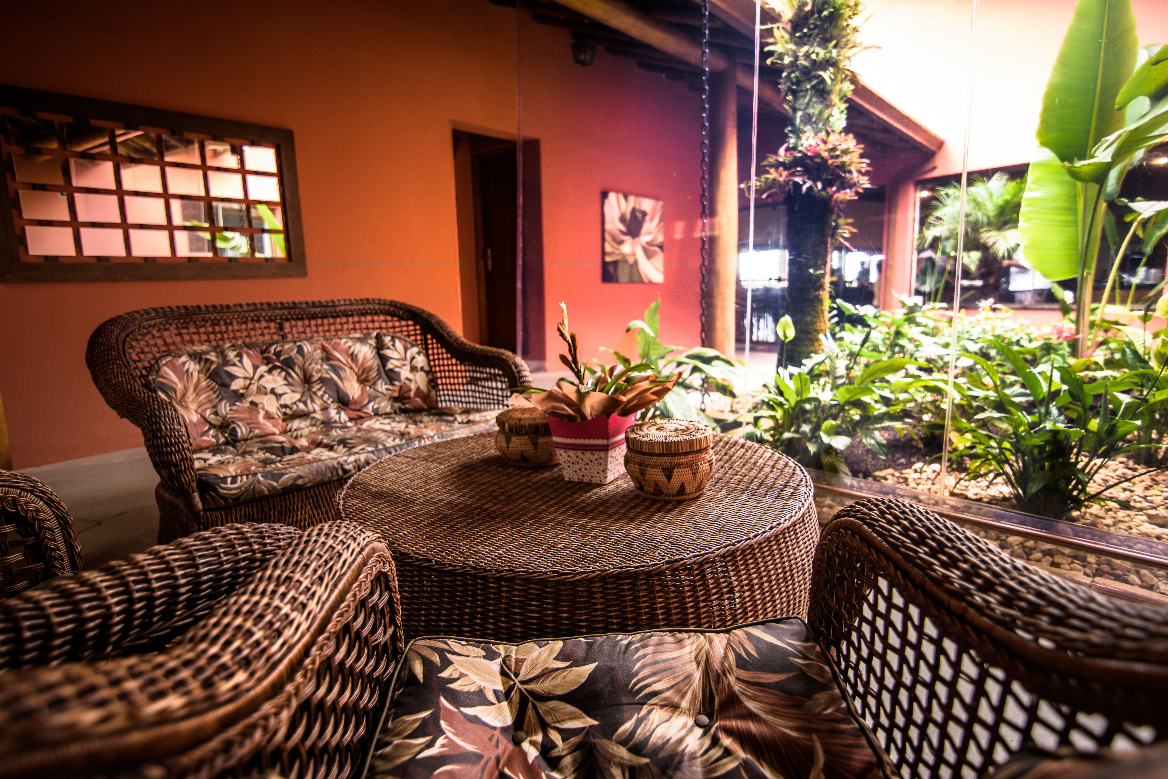 Conjunto de móveis de vime com uma mesa redonda, um sofá e duas poltronas, dispostos num espaço com vista para um jardim.