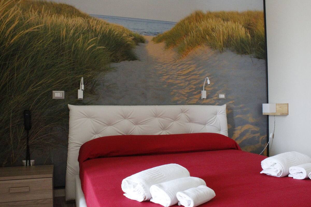 Quarto de hotel com cama, roupa de cama vermelha e um mural com grama e água.