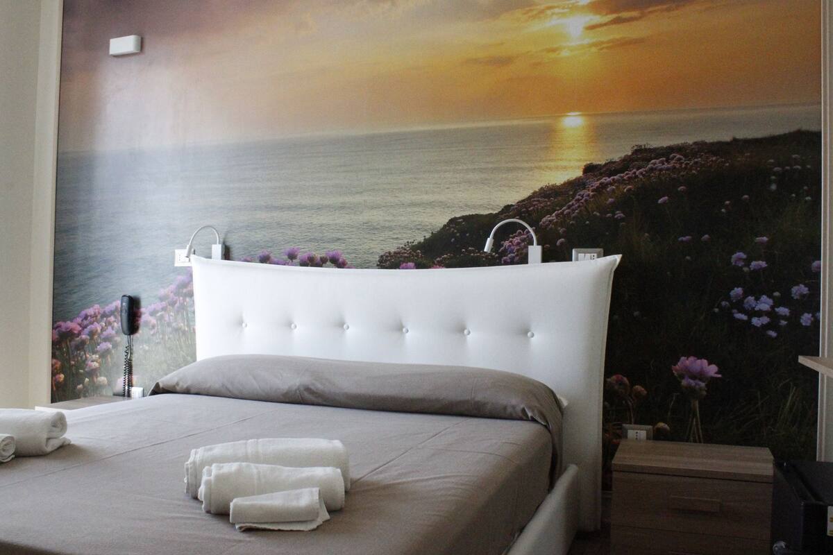 Quarto com uma cama grande, cabeceira branca e um mural de um pôr do sol sobre o oceano.