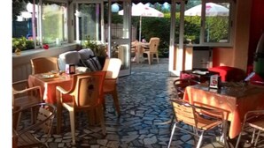 Uma área de estar coberta ao ar livre, com mesas e cadeiras, piso de azulejos e vista para o jardim através de portas de vidro.