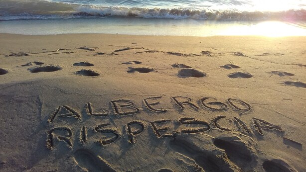 Pegadas na praia com a frase 'ALBERGO RISPECIA' escrita na areia.