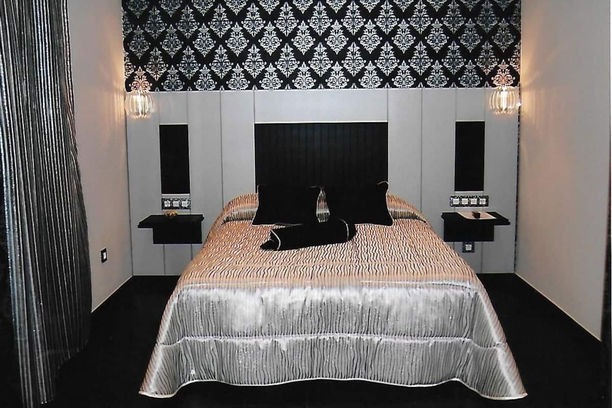 Quarto moderno com papel de parede estampado, uma cama grande com colcha metálica, duas mesas de cabeceira com luminárias e cabeceira preta.