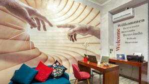 Uma área de recepção com design moderno, apresentando um mural grande na parede, um balcão de madeira, um sofá azul com almofadas vermelhas e um vaso de planta.