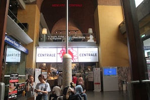 Um espaço amplo e interno, com teto alto e uma coluna central, apresentando uma placa com 'CENTRALE' e um quiosque de informações digital.