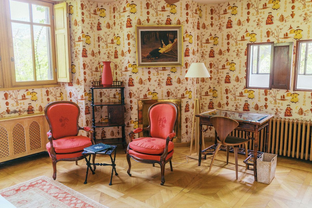 Quarto com papel de parede estampado, duas cadeiras vermelhas, uma escrivaninha de madeira, um radiador e uma janela.
