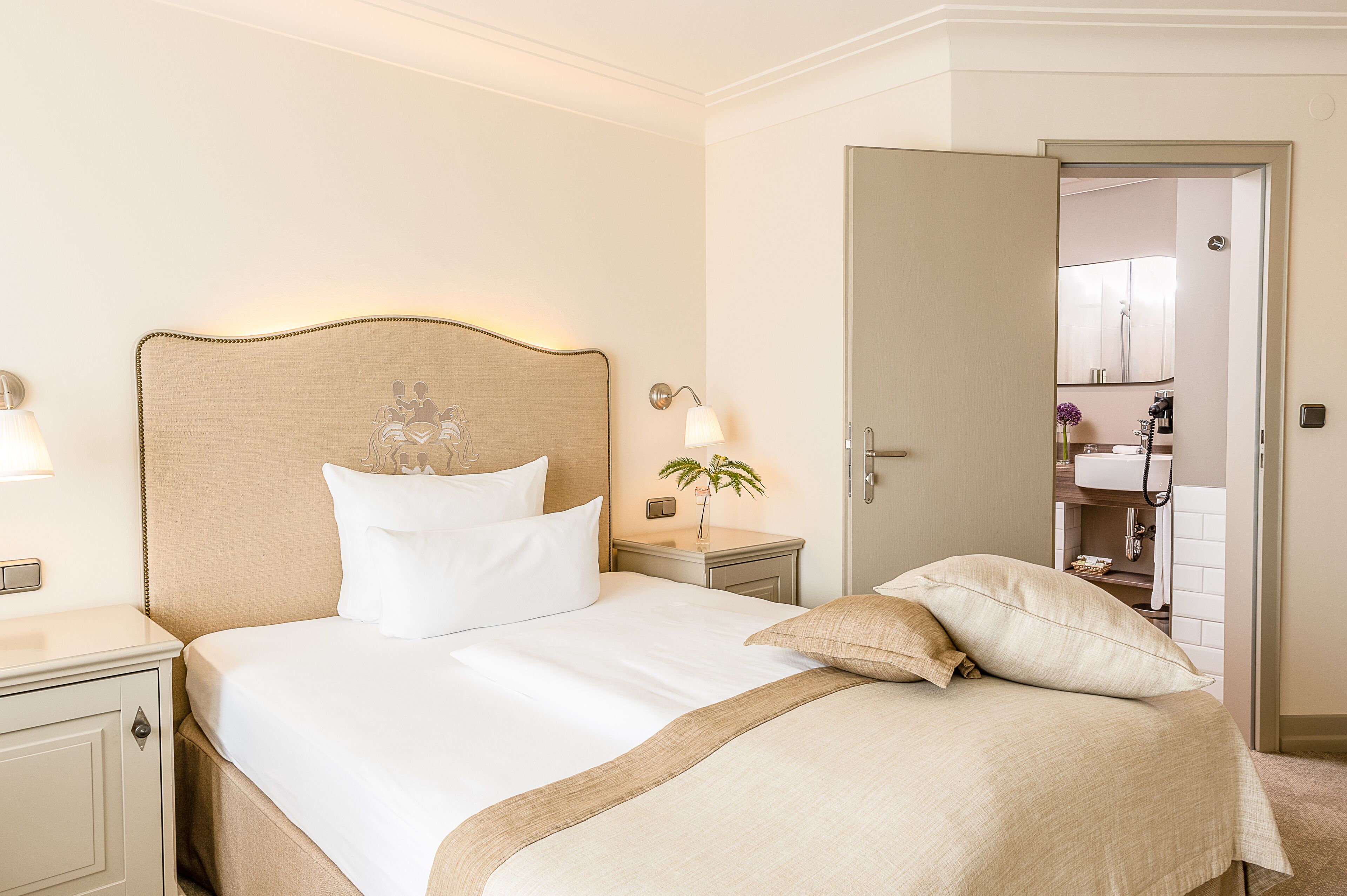 Uma cama bem arrumada com lençóis brancos e cabeceira bege, uma luminária de cabeceira e uma mesinha com um vaso de flores.