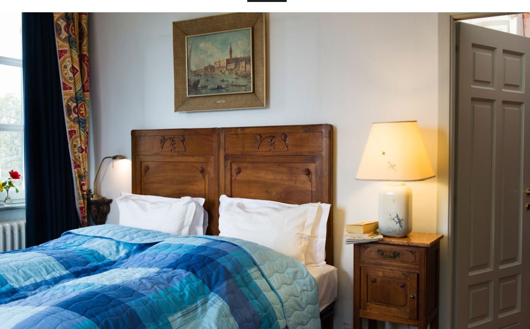 Uma cama bem arrumada com um edredom azul e verde, uma cabeceira de madeira, um abajur, uma pequena mesa de cabeceira de madeira e um quadro na parede.