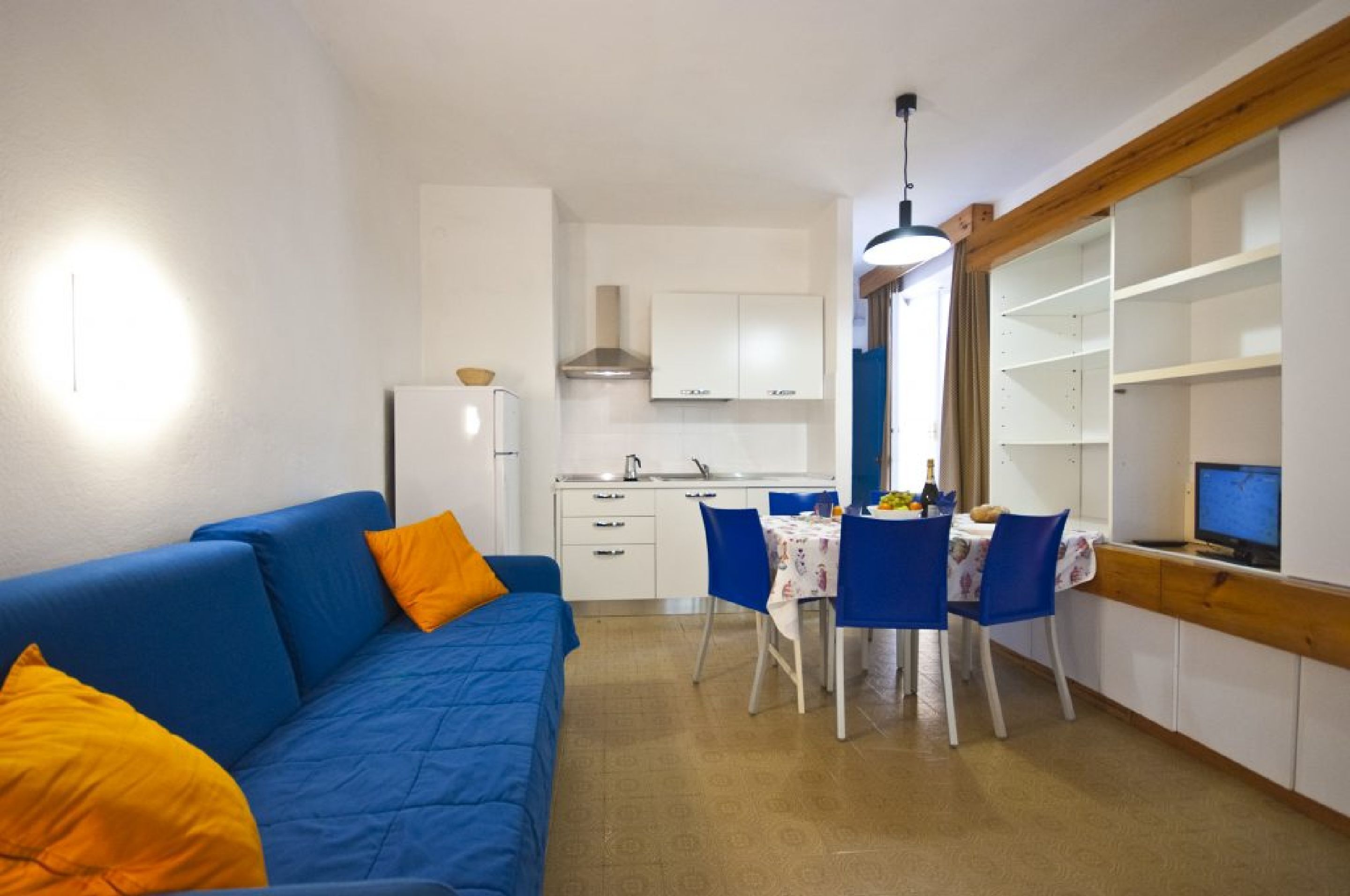 Um espaço de estar compacto com um sofá azul, uma mesa de jantar com cadeiras, uma área de cozinha e um computador em uma escrivaninha.