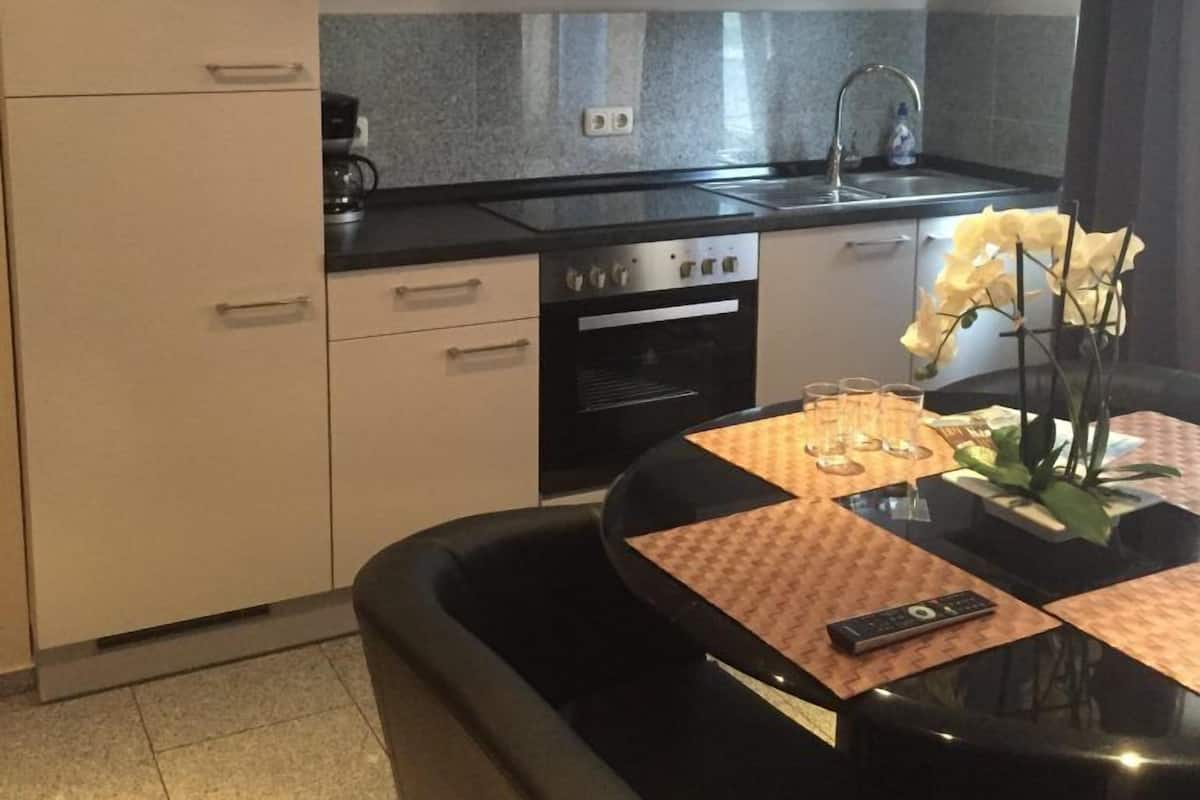 Uma cozinha compacta com fogão, forno e armários modernos. Há uma área de refeições com uma mesa posta para dois, com um vaso de flores.