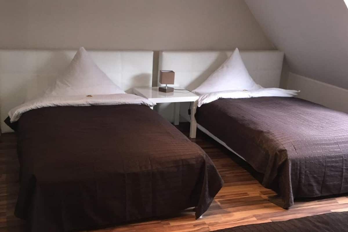 Duas camas de solteiro com almofadas brancas e colchas castanhas em um quarto com teto inclinado e piso de madeira.