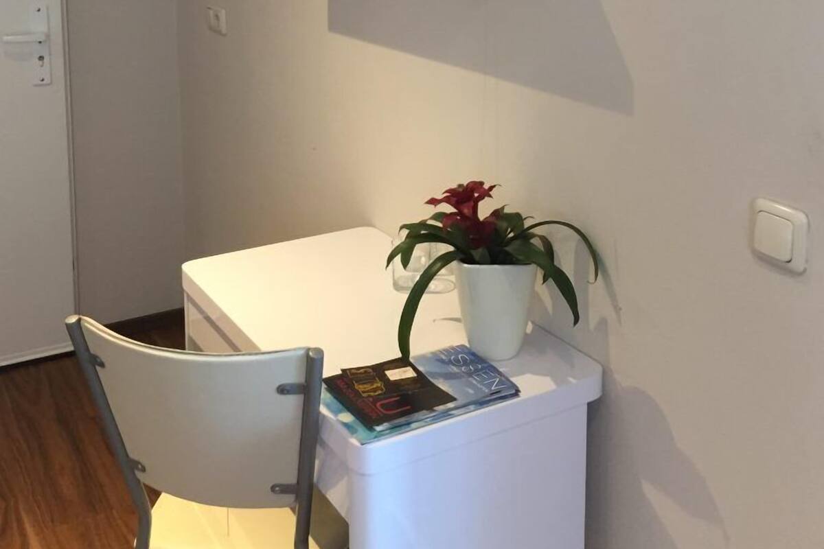 Uma secretária pequena e branca com uma cadeira, uma TV montada, uma prateleira com um comando remoto, uma planta em vaso e alguns livros.