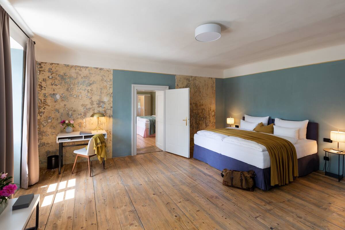 Quarto de hotel com piso de madeira, cama com roupa de cama branca e marrom, escrivaninha com luminária e vista para outro quarto por uma porta aberta.