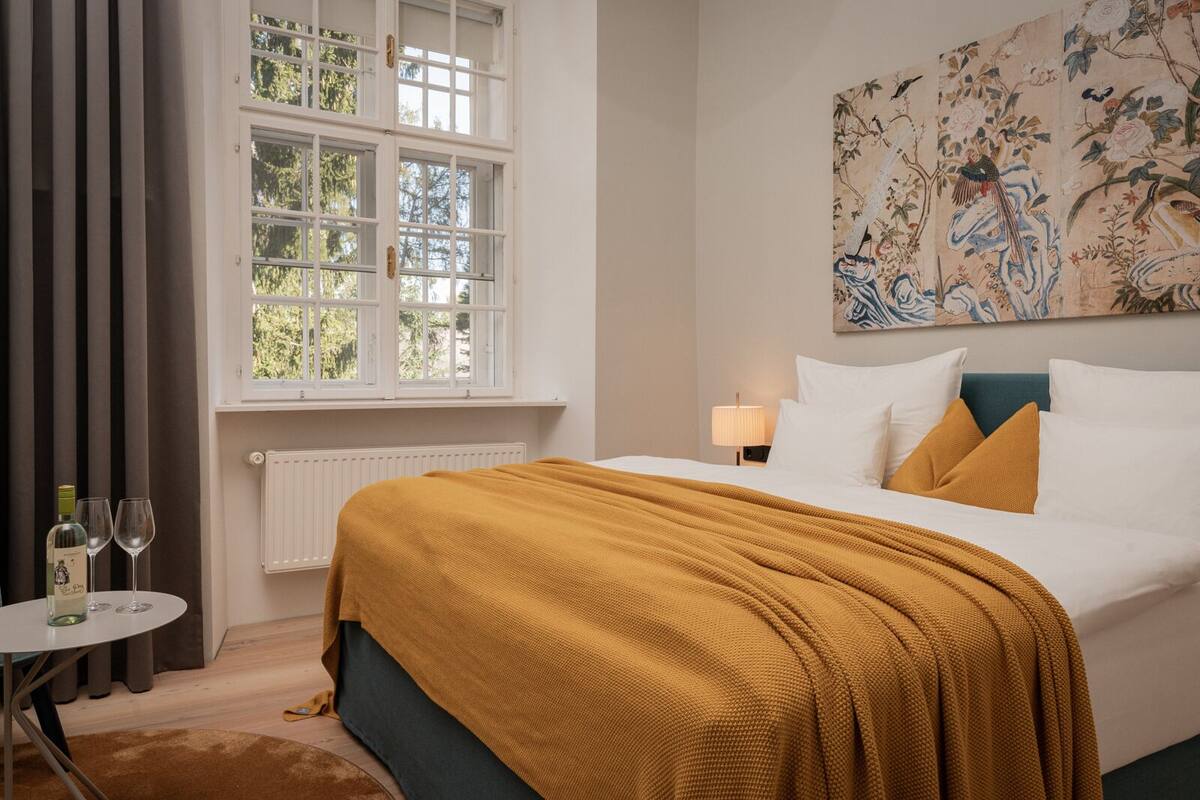 Quarto de hotel com cama, janela com cortinas, mesa com copos e uma garrafa, e um quadro na parede.
