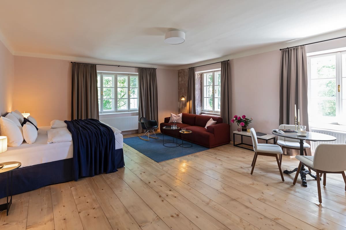 Quarto de hotel com uma cama, um sofá, uma mesa de jantar e uma cadeira. Há janelas com cortinas, um abajur e um vaso com flores.
