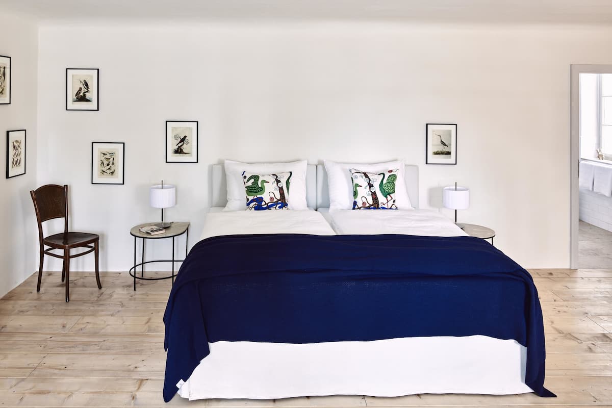 Um quarto com uma cama grande, duas almofadas com desenhos de pássaros, uma cadeira de madeira, duas mesas de cabeceira com abajures e quadros nas paredes.