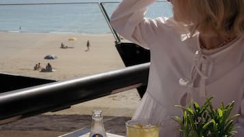 Mesa com bebida, garrafa e chávena, com vista para uma praia com pessoas e guarda-sóis.