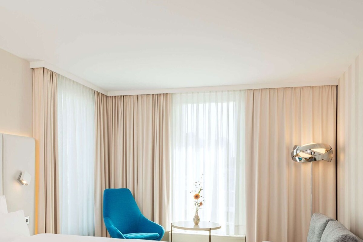 Quarto de hotel com uma cama, uma cadeira azul, uma mesinha com um vaso e uma televisão presa na parede.