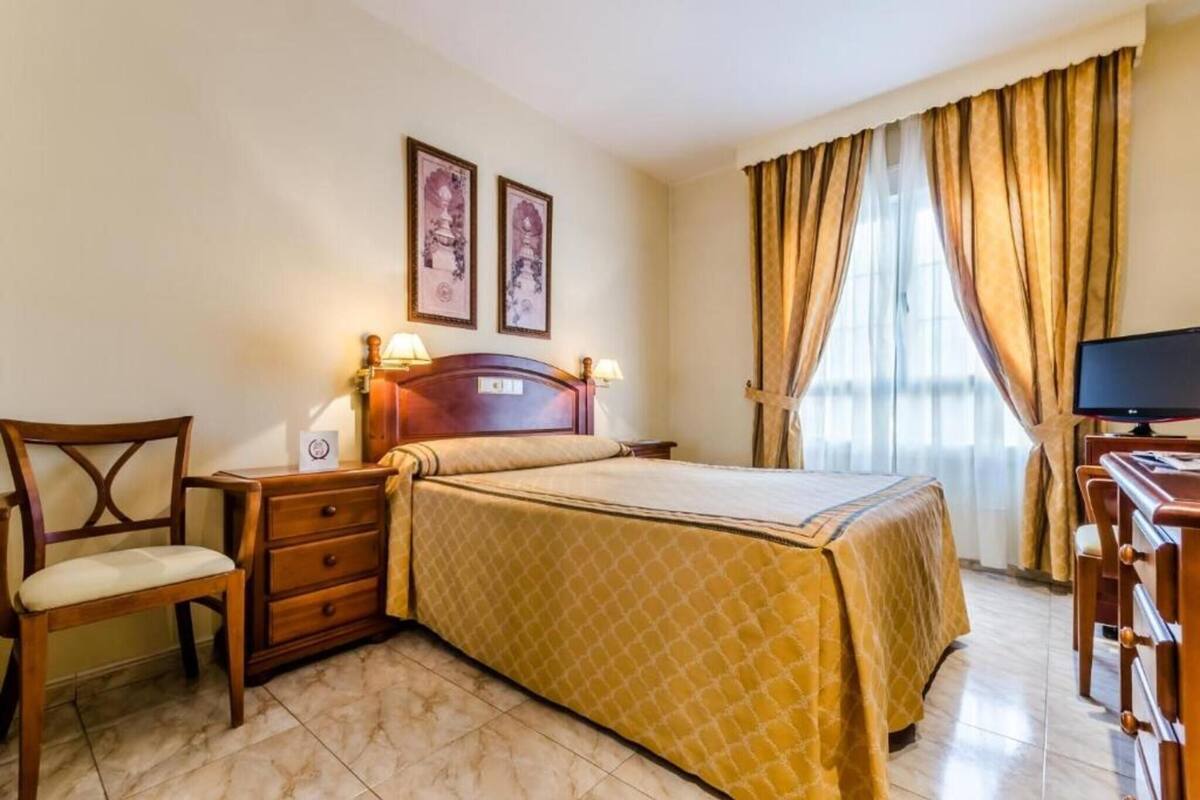Quarto de hotel com cama, escrivaninha, cadeira, televisão e duas molduras na parede.