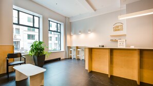 Uma área de recepção moderna com balcão, dois bancos altos, uma planta em vaso, uma mesa branca e grandes janelas.
