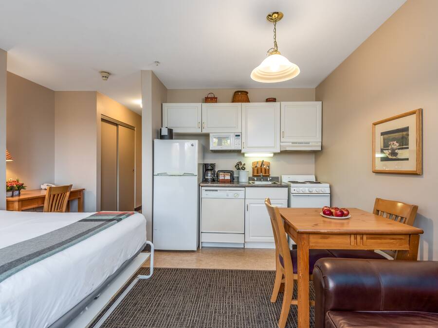 Quarto de hotel compacto com uma cama, pequeno espaço de cozinha, área de jantar com mesa e cadeiras, e uma única poltrona de couro marrom.