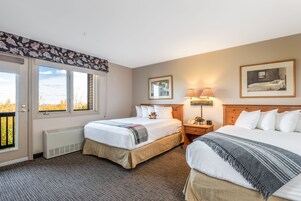 Quarto de hotel com duas camas, uma janela grande, um radiador, uma mesa de cabeceira com luminária e um quadro na parede.