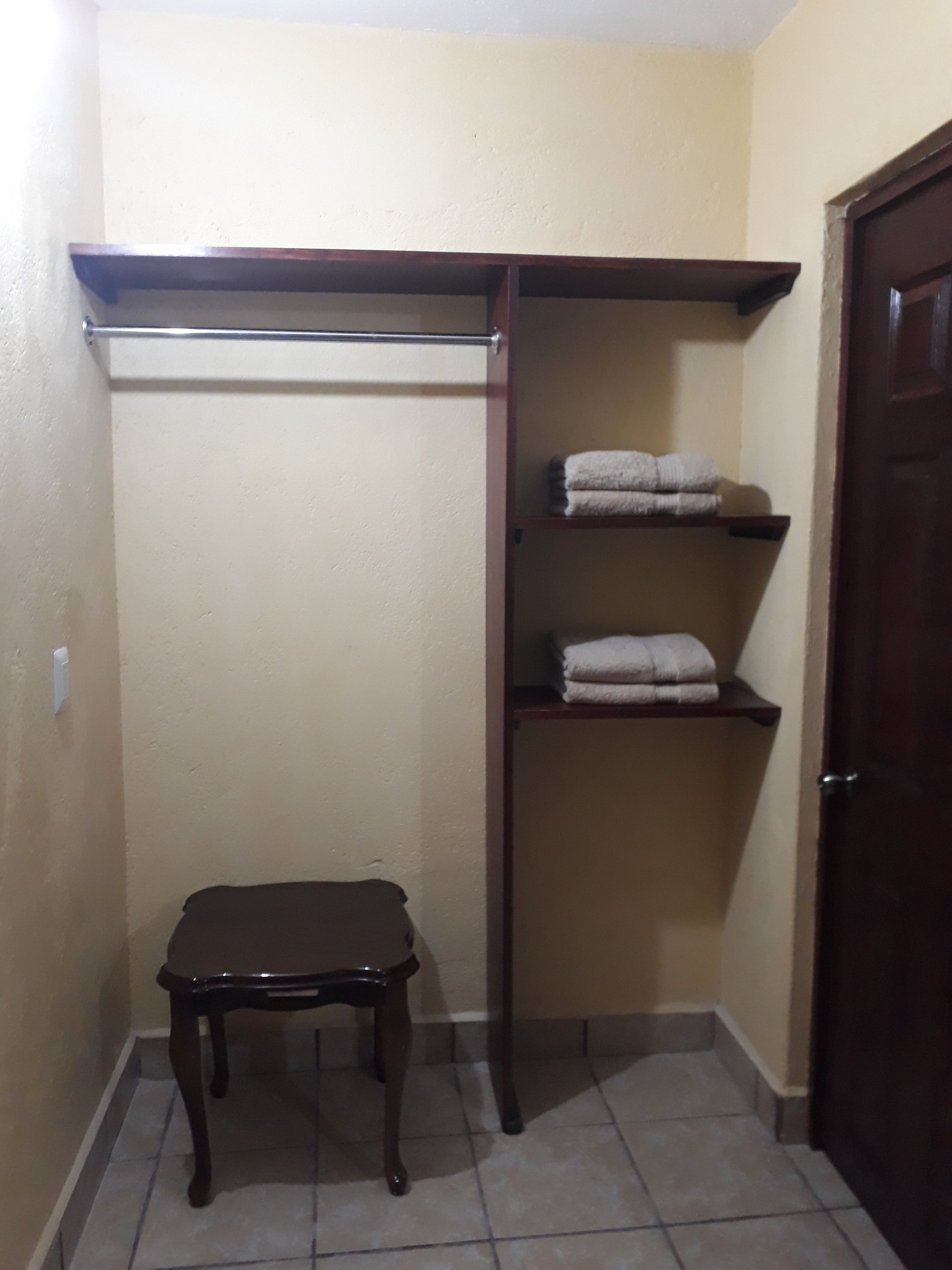 Um quarto pequeno com uma cadeira de madeira, uma prateleira com toalhas dobradas e uma porta.