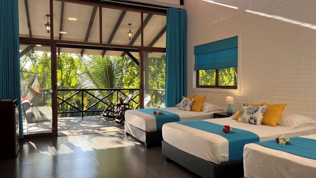 Quarto de hotel com duas camas, varanda com vista para área verde e parede de tijolos brancos.