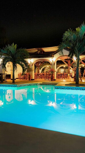 Piscina com borda azulejada, rodeada por palmeiras e um edifício com entrada em arco.