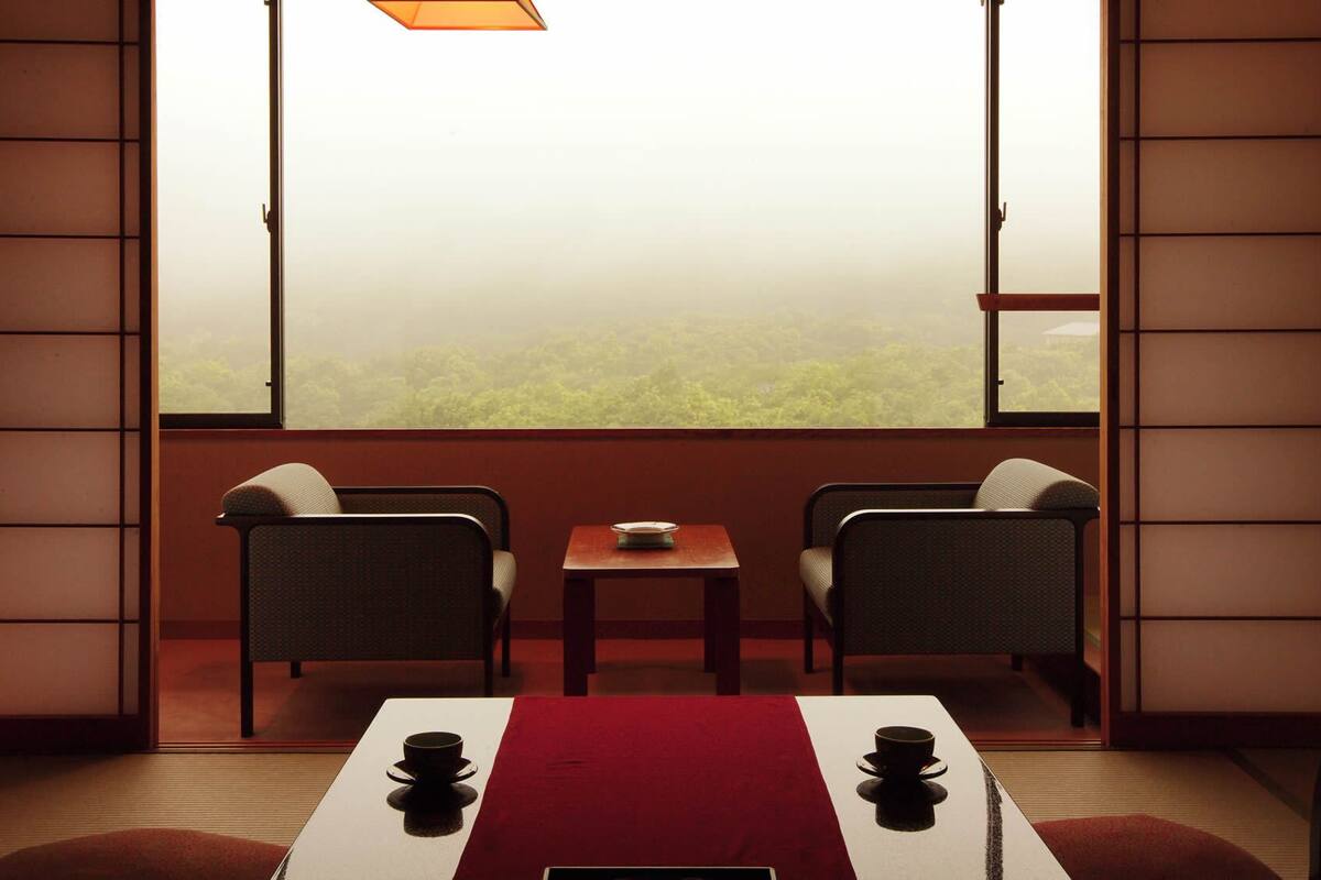 Quarto com piso de esteira de tatame, mesa baixa com um tapete vermelho, duas cadeiras e vista para uma floresta através de grandes janelas.