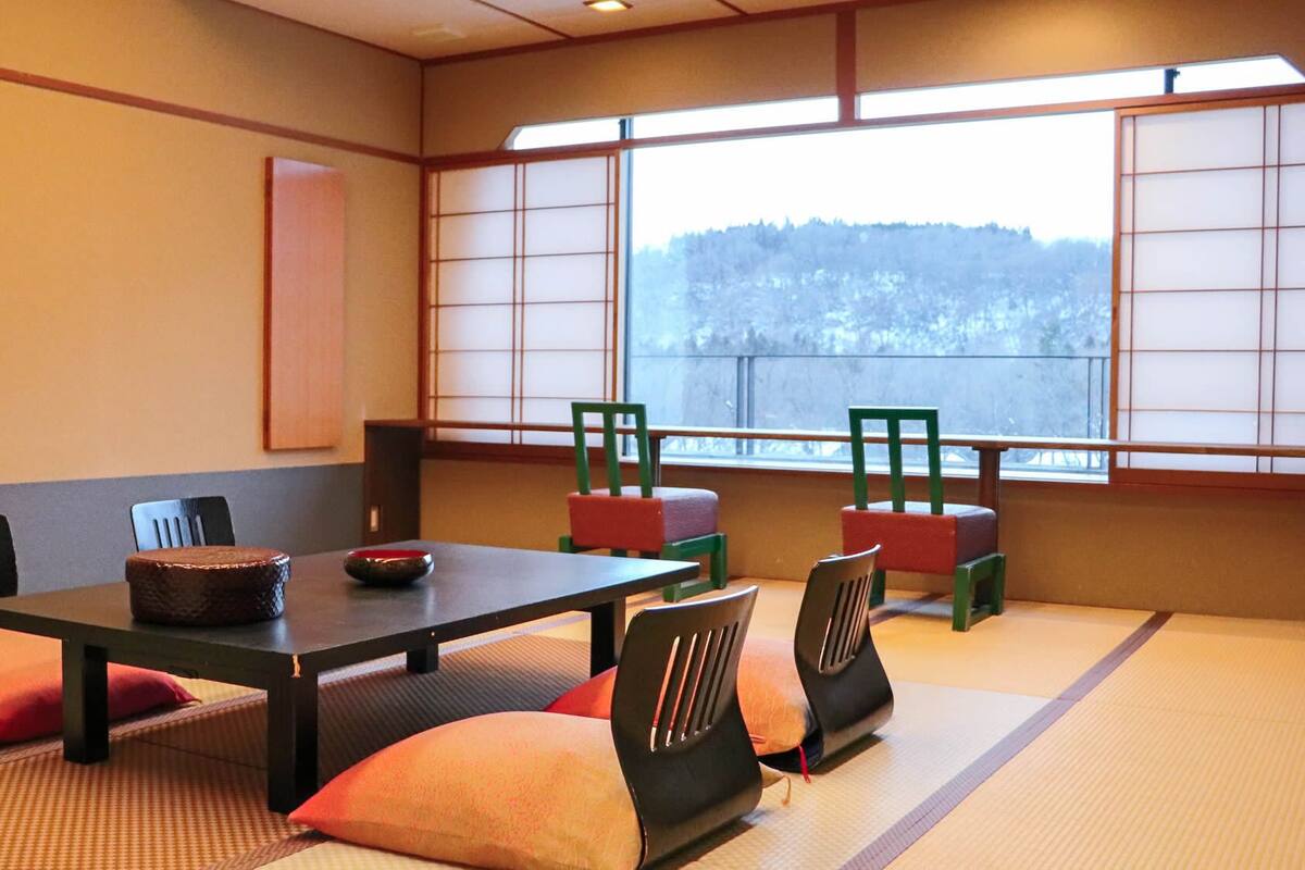 Um espaço com tatames, uma mesa baixa, cadeiras e vista através de grandes janelas.
