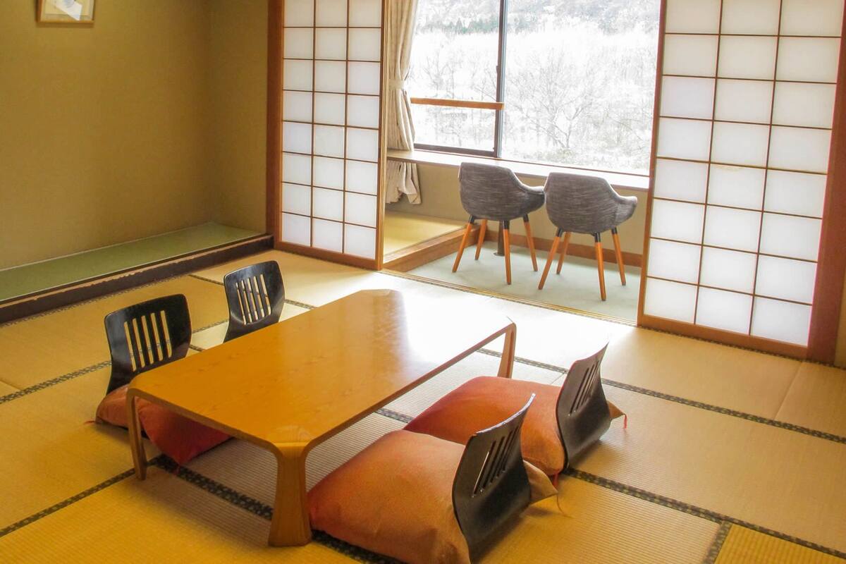 Um espaço com tatames, uma mesa baixa e cadeiras, uma porta de correr para uma varanda com cadeiras, e vista para uma paisagem nevada.