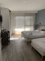 Quarto de hotel com duas camas, piso de madeira, uma janela grande com cortinas e uma pequena mesa com chaleira e garrafa.