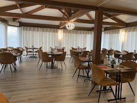 Uma ampla sala de jantar com vigas de madeira, mesas postas com cadeiras e grandes janelas com cortinas.