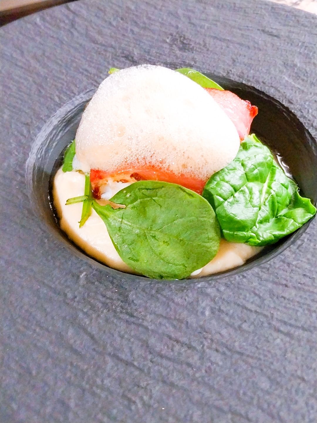 Um prato escuro com um item branco esférico, folhas verdes e uma fatia de carne vermelha.