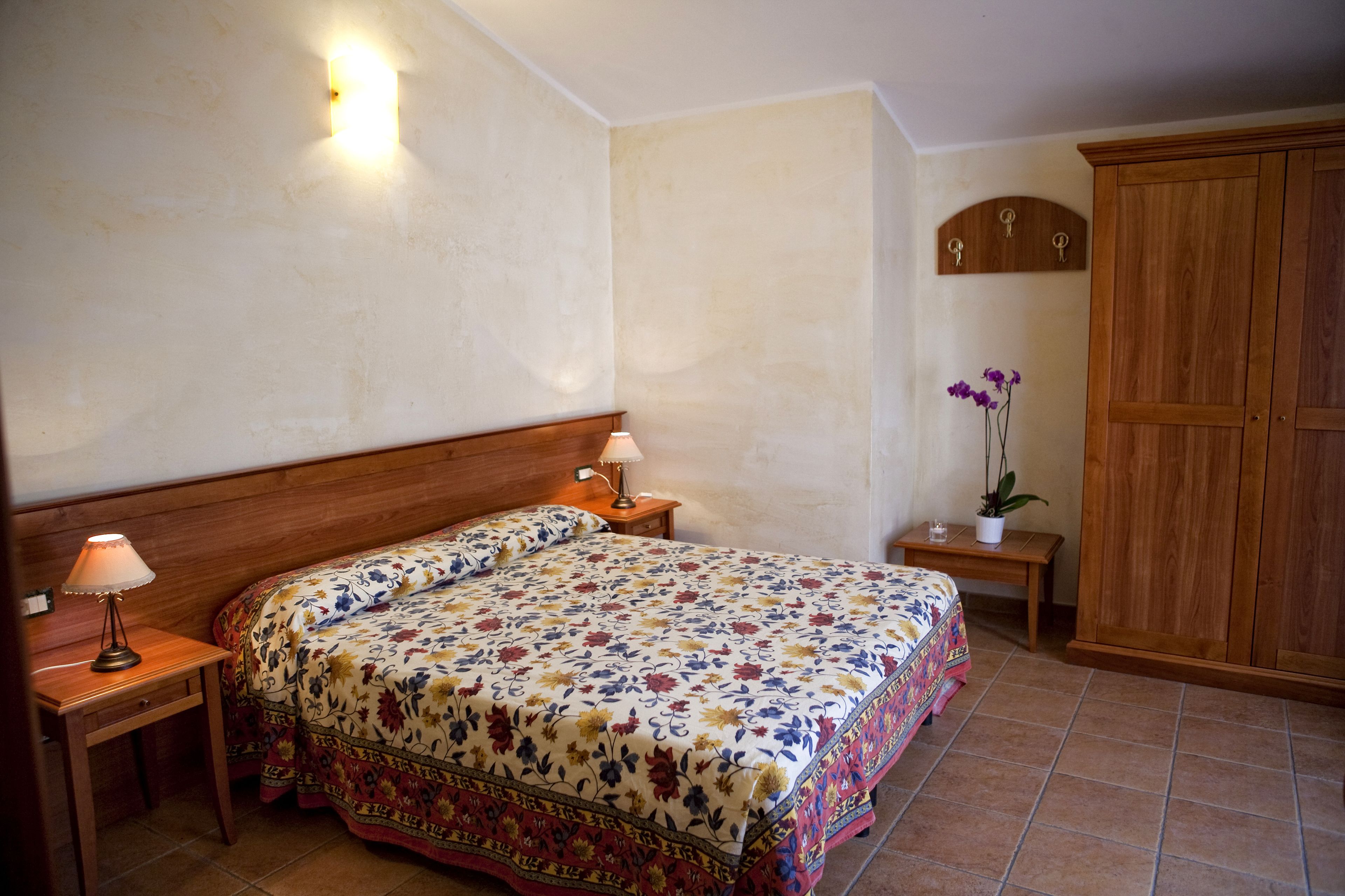 Quarto com uma cama grande, duas mesas de cabeceira com abajures, um guarda-roupa de madeira e uma prateleira na parede com um vaso de planta.