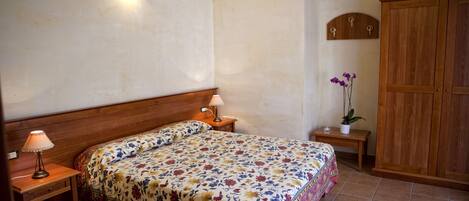 Quarto com uma cama grande, duas mesas de cabeceira com abajures, um guarda-roupa de madeira e uma prateleira na parede com um vaso de planta.