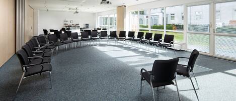 Uma sala de conferência com disposição semicircular de cadeiras, amplas janelas e um projetor no teto.